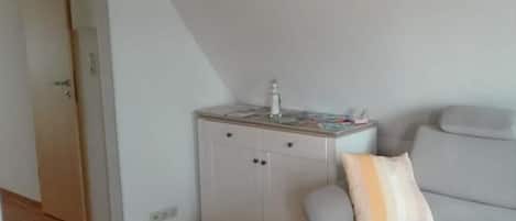 Quarto com sofá cinzento, armário branco com gavetas e uma pequena mesa com abajur e livros.