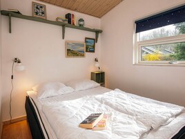 Um quarto com uma cama, uma mesa de cabeceira, uma estante com livros e um candeeiro, um quadro, e uma janela com vista para árvores e painéis solares.