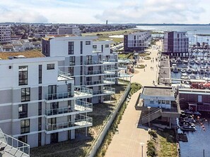 Um edifício moderno de vários andares com varandas, uma marina com barcos e vista para o litoral.