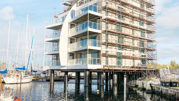 Edifício de vários andares em construção, com vista para o cais, sustentado por pilares.