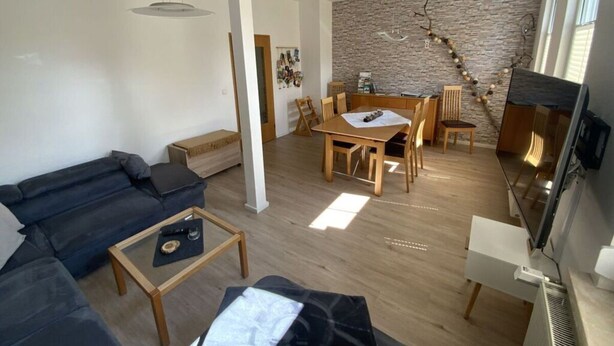 Uma sala de estar moderna com um sofá escuro, uma mesa de centro de madeira e um espaço de jantar com mesa e cadeiras de madeira.