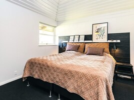 Quarto com cama, mesa de cabeceira, prateleira fixada na parede com quadros e janela.