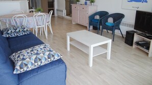 Uma sala de estar moderna com sofá, mesa de jantar com cadeiras, rack de TV com televisão de tela plana e área de cozinha ao fundo.