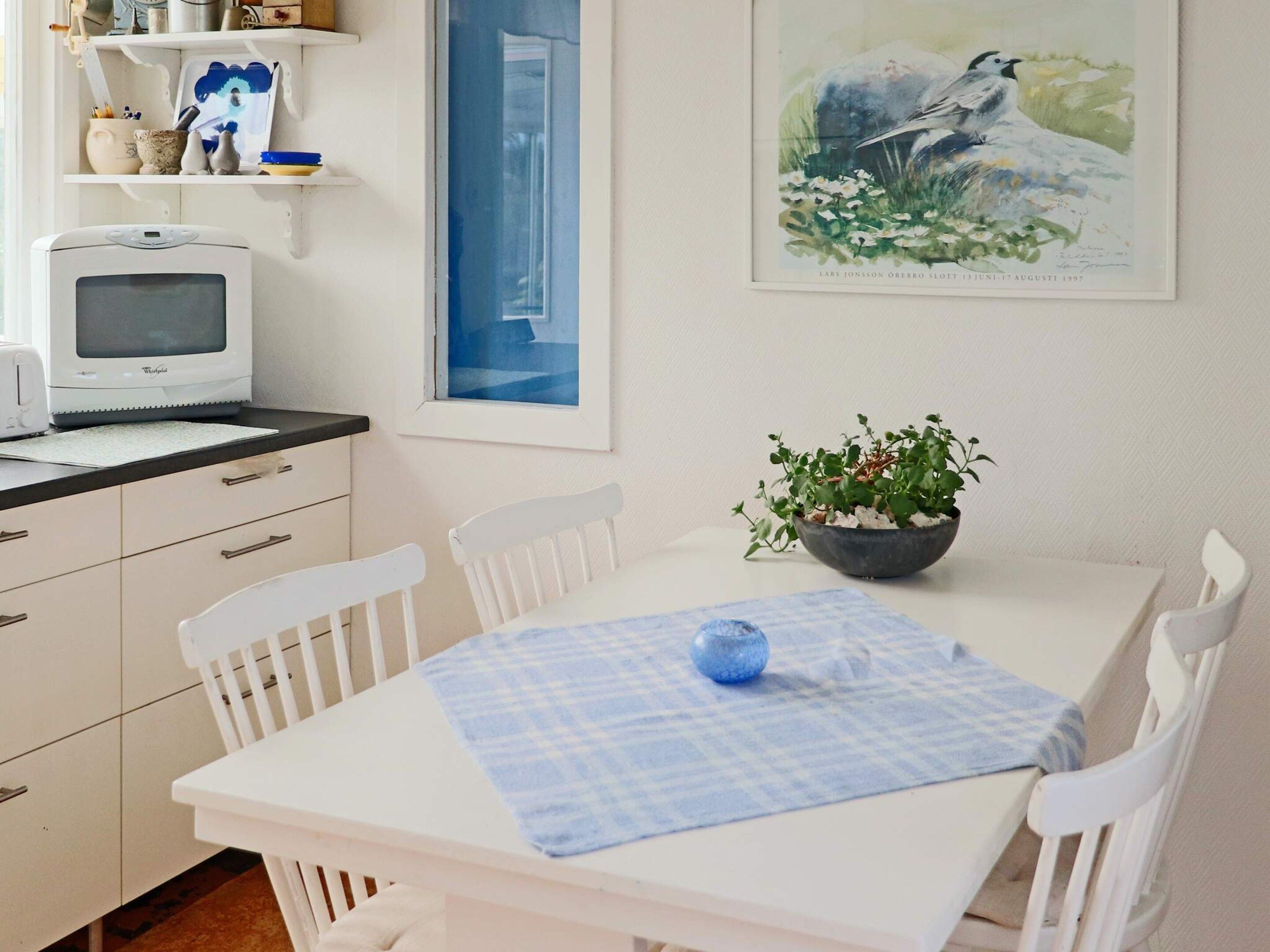 Mesa de jantar branca com toalha xadrez azul, uma tigela com plantas verdes e um quadro emoldurado de um pássaro.