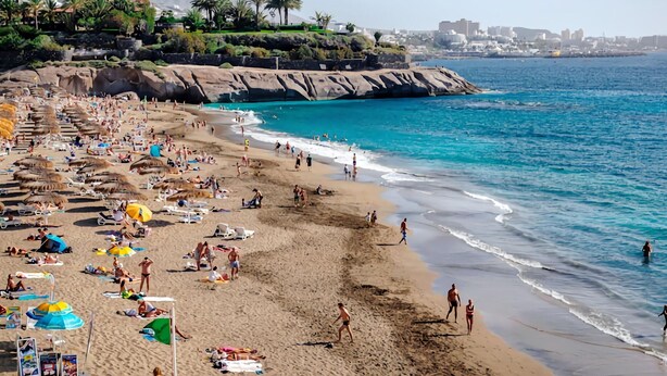 Uma praia com pessoas se banhando ao sol e nadando, um trecho de costa rochosa e edifícios ao fundo.