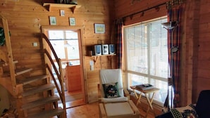 Interior de cabana de madeira com escada, uma cadeira com almofada, mesa pequena e janela com persianas.
