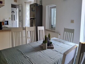 Uma área de refeições com uma mesa posta para dois, cadeiras brancas, uma pequena planta em vaso e uma janela com persianas.