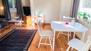 Um espaço de estar compacto com uma mesa de jantar, cadeiras brancas, uma pequena escrivaninha, uma televisão e uma janela com cortinas.