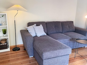 Um sofá modular cinza com duas almofadas combinando, uma pequena mesa redonda, um abajur e uma estante branca com objetos decorativos.