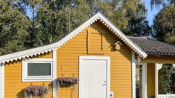 Um chalé de madeira amarela com porta e janela brancas, um pequeno alpendre e um jardim com flores.