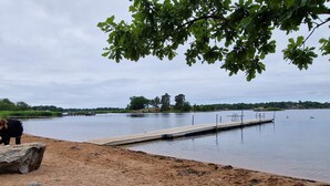Vista para o lago com um píer que se estende sobre a água, cercado por árvores e uma margem arenosa.
