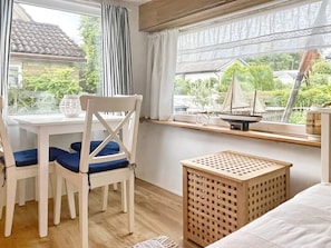 Quarto com piso de madeira, uma cadeira branca com almofada azul, uma pequena mesa de madeira e uma janela com vista para área verde e um veleiro.