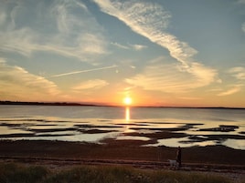 Pôr do sol sobre a água, com uma pessoa e um cão a caminhar por um trilho.