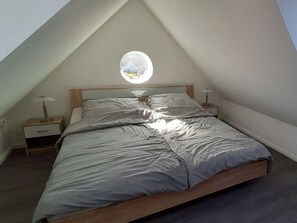 Quarto com teto inclinado, um claraboia circular, cama com roupa de cama cinzenta e duas mesas de cabeceira com abajures.