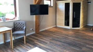Quarto com teto de madeira, televisão de tela plana na parede, poltrona cinza estofada, mesa de madeira pequena e porta de vidro deslizante para uma varanda.