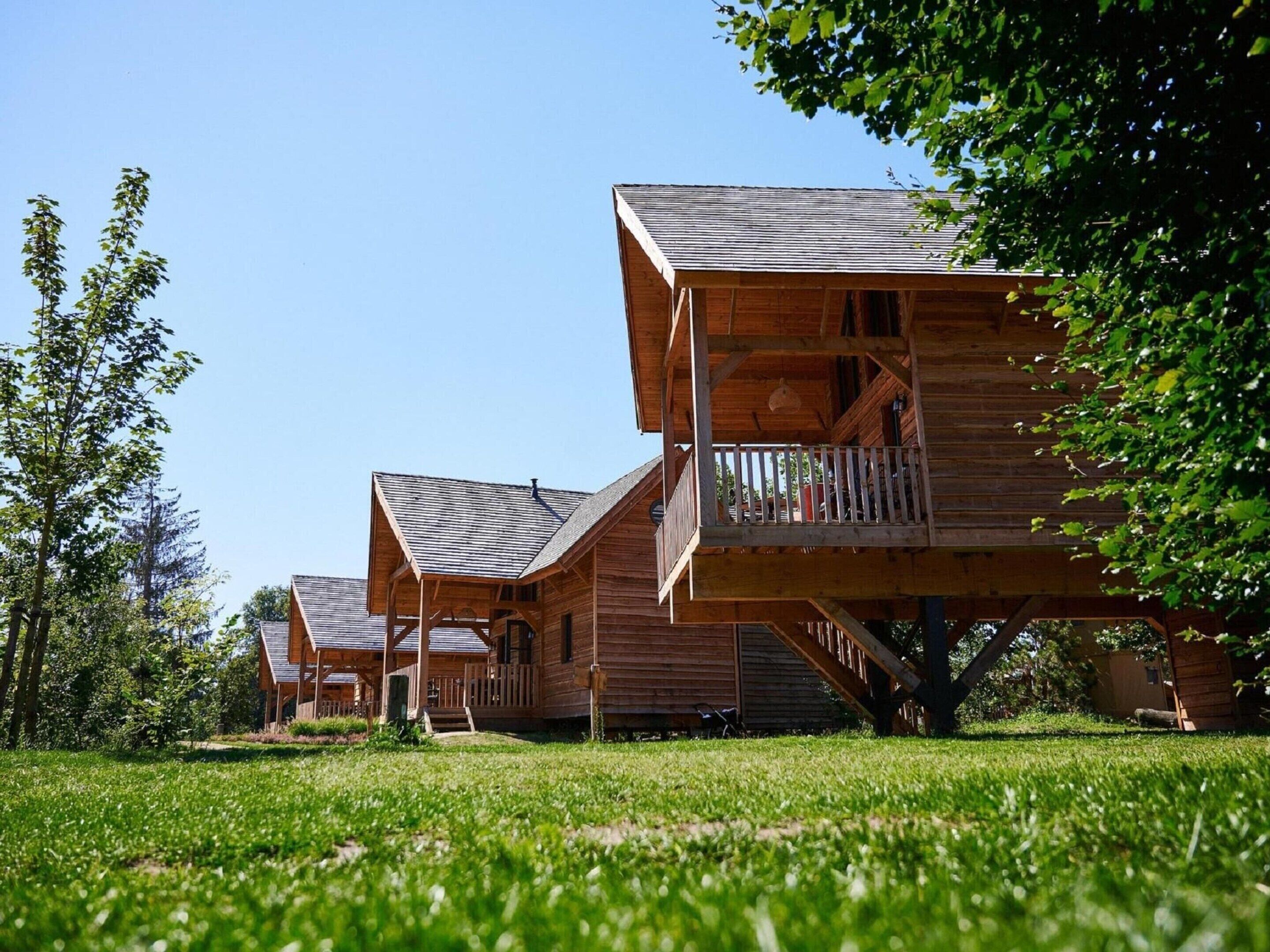 Uma fileira de cabanas de madeira com plataformas e guarda-corpos elevados, localizada numa área ajardinada com árvores.