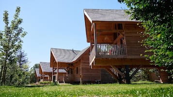 Uma fileira de cabanas de madeira com plataformas e guarda-corpos elevados, localizada numa área ajardinada com árvores.