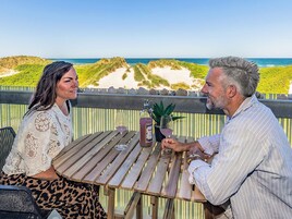Duas pessoas sentadas a uma mesa de madeira, com vista para a praia e dunas ao fundo.