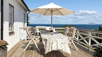 Um deck de madeira com uma mesa, cadeiras e um guarda-sol branco, com vista para o mar.