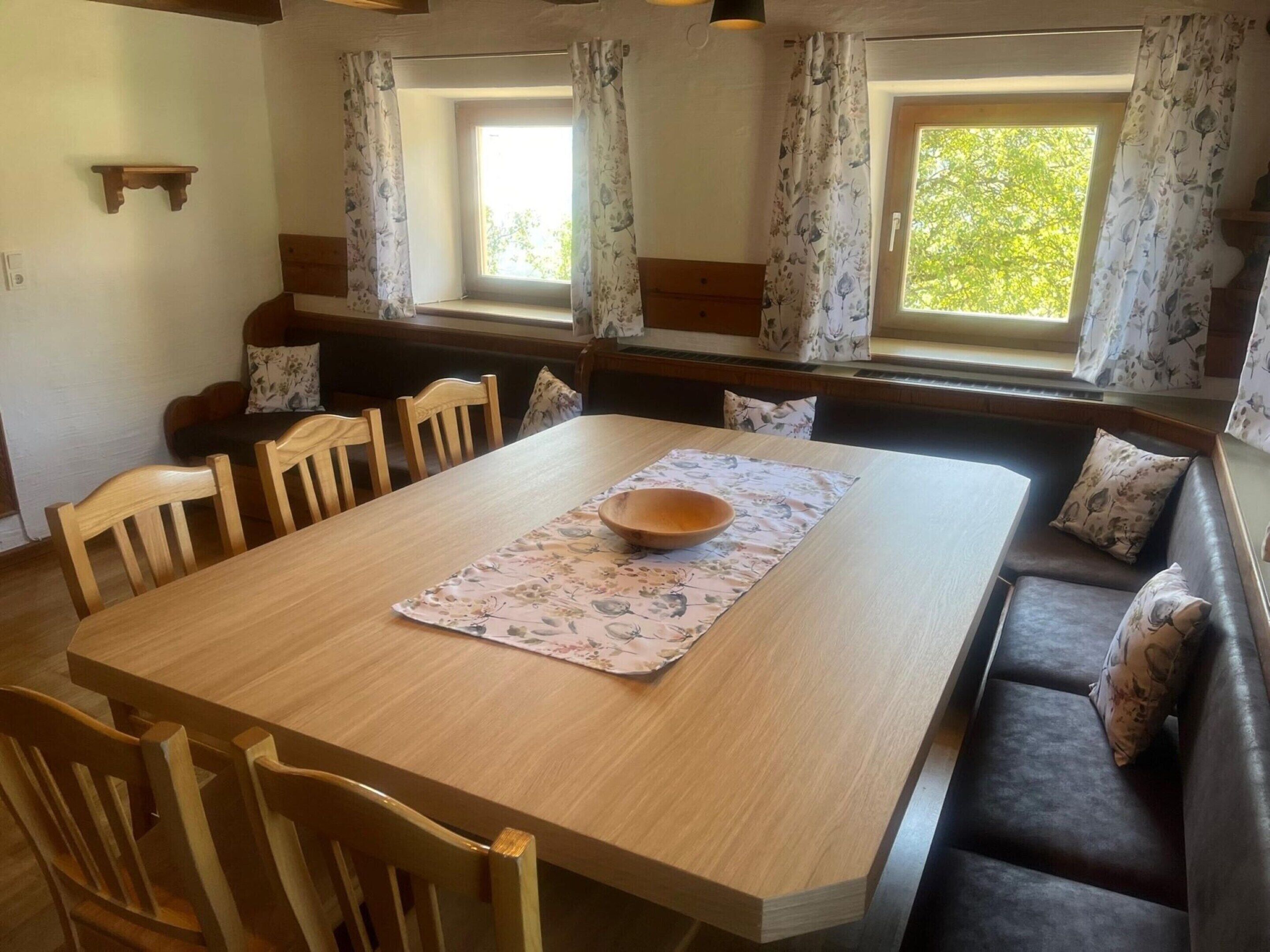 Uma área de refeições com uma mesa de madeira, um jogo americano floral e uma tigela. Há cadeiras de madeira e um sofá escuro com almofadas florais. Duas janelas com cortinas florais proporcionam luz natural.