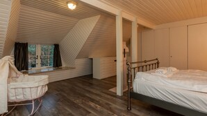 Quarto com teto inclinado, uma cama, cômoda e janela com cortinas.