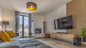 Uma sala de estar moderna com um sofá modular cinza, uma TV de tela plana fixada em uma parede de madeira e uma janela grande com cortinas.