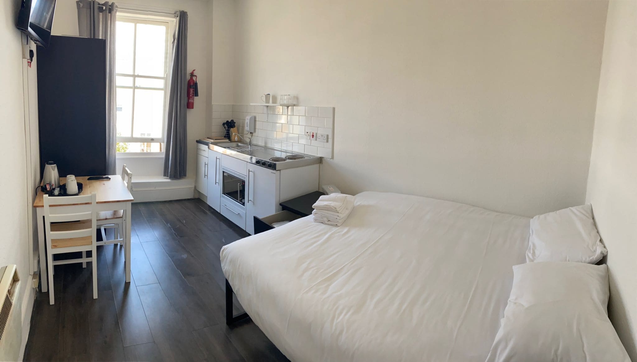 Quarto de hotel com cama, escrivaninha com cadeira, cozinha compacta e janela com cortinas.