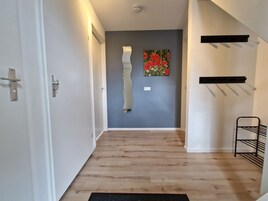 Um corredor com piso de madeira, porta à esquerda, prateleira na parede à direita e um quadro com flores vermelhas na parede.