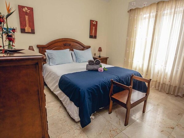 Quarto bem arrumado com cama, cadeira de madeira, mesa de cabeceira e escrivaninha.