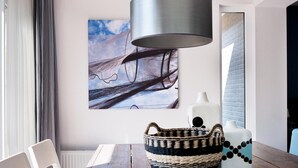 Uma sala de jantar com uma mesa de madeira, um cesto trançado, um vaso branco e um quadro na parede.