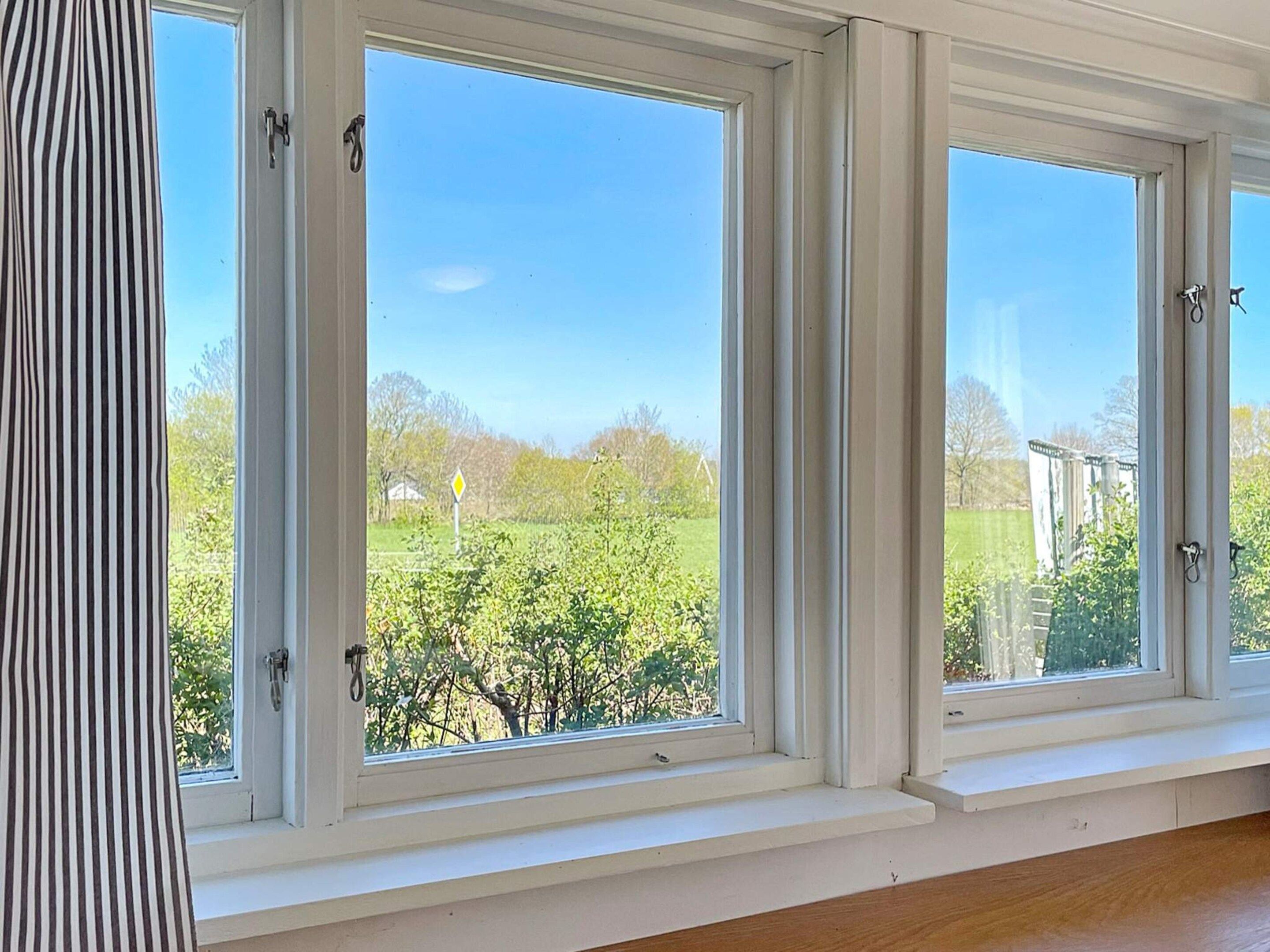 Quarto com três grandes janelas, cada uma com moldura branca e vista para uma paisagem verde com árvores e céu limpo.