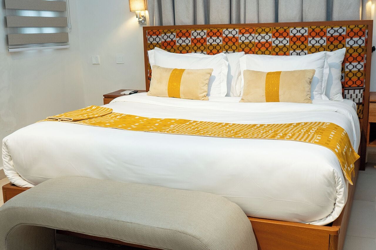 Uma cama bem arrumada com cabeceira estampada, mesa de cabeceira com luminária e um banco aos pés da cama.