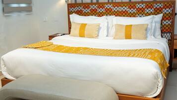 Uma cama bem arrumada com cabeceira estampada, mesa de cabeceira com luminária e um banco aos pés da cama.