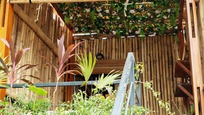 Estrutura de bambu com telhado de palha, rodeada por diversas plantas e uma escadaria de madeira.