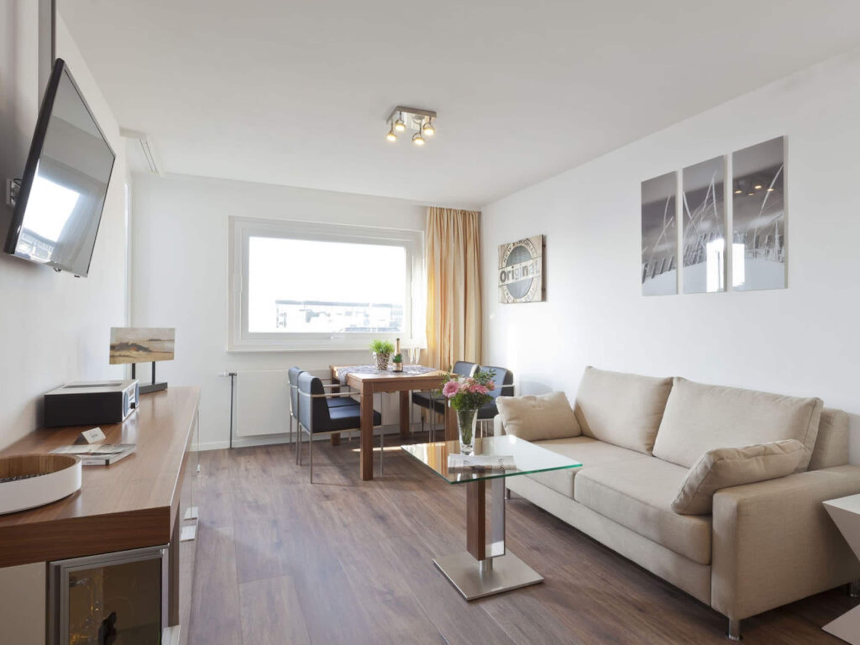 Uma sala de estar moderna com uma mesa de centro de vidro, um sofá bege, uma área de jantar com mesa e cadeiras, uma televisão de tela plana na parede e uma grande janela com cortinas.