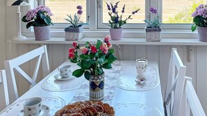 Uma mesa de jantar com um arranjo floral, uma cesta de pastelaria e vista para um campo pela janela.