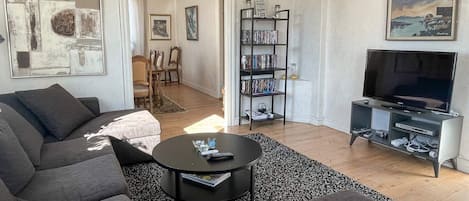 Uma sala de estar com sofá, mesa de centro, estante de livros, televisão e um quadro na parede.