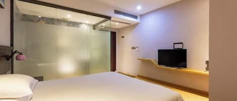 Quarto de hotel moderno com uma cama grande, uma divisória de vidro, uma televisão sobre uma estante de madeira e uma luminária fixada na parede.