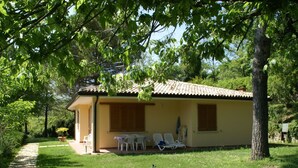 Um pequeno edifício térreo com telhado de telhas e varanda coberta. Há várias cadeiras de plástico branco e uma mesa na varanda.