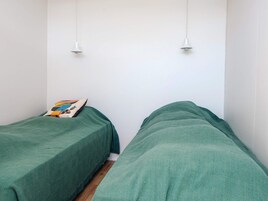 Quarto pequeno e limpo com duas camas de solteiro, cada uma com colcha verde e almofada colorida. Há duas luminárias pendentes brancas no teto.