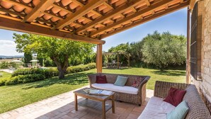 Um pátio externo com móveis de vime, uma pérgola de madeira e vista para um jardim exuberante e colinas ao longe.