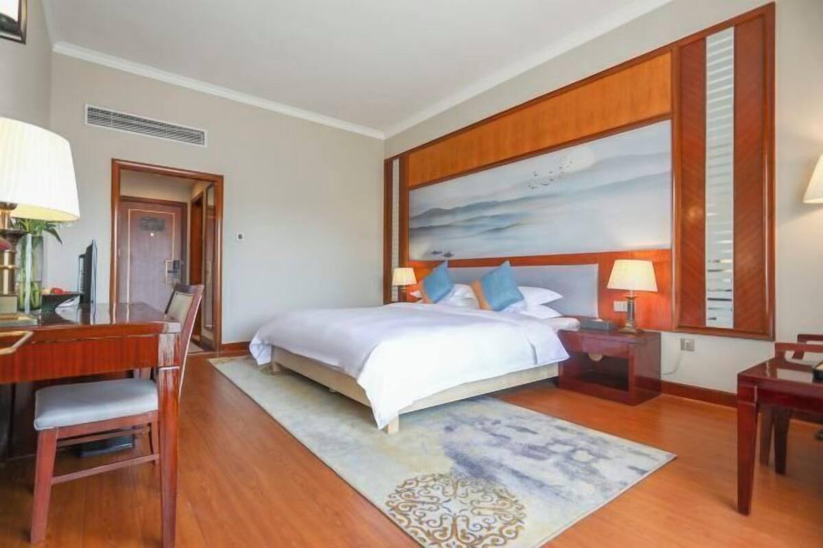 Quarto de hotel com cama, escrivaninha, cadeira, mesa de cabeceira e uma pintura grande na parede.