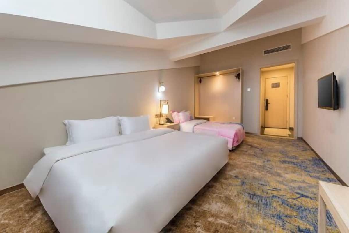 Quarto de hotel com uma cama grande, uma mesa de cabeceira com abajur, uma televisão na parede e uma porta que dá para outro quarto.