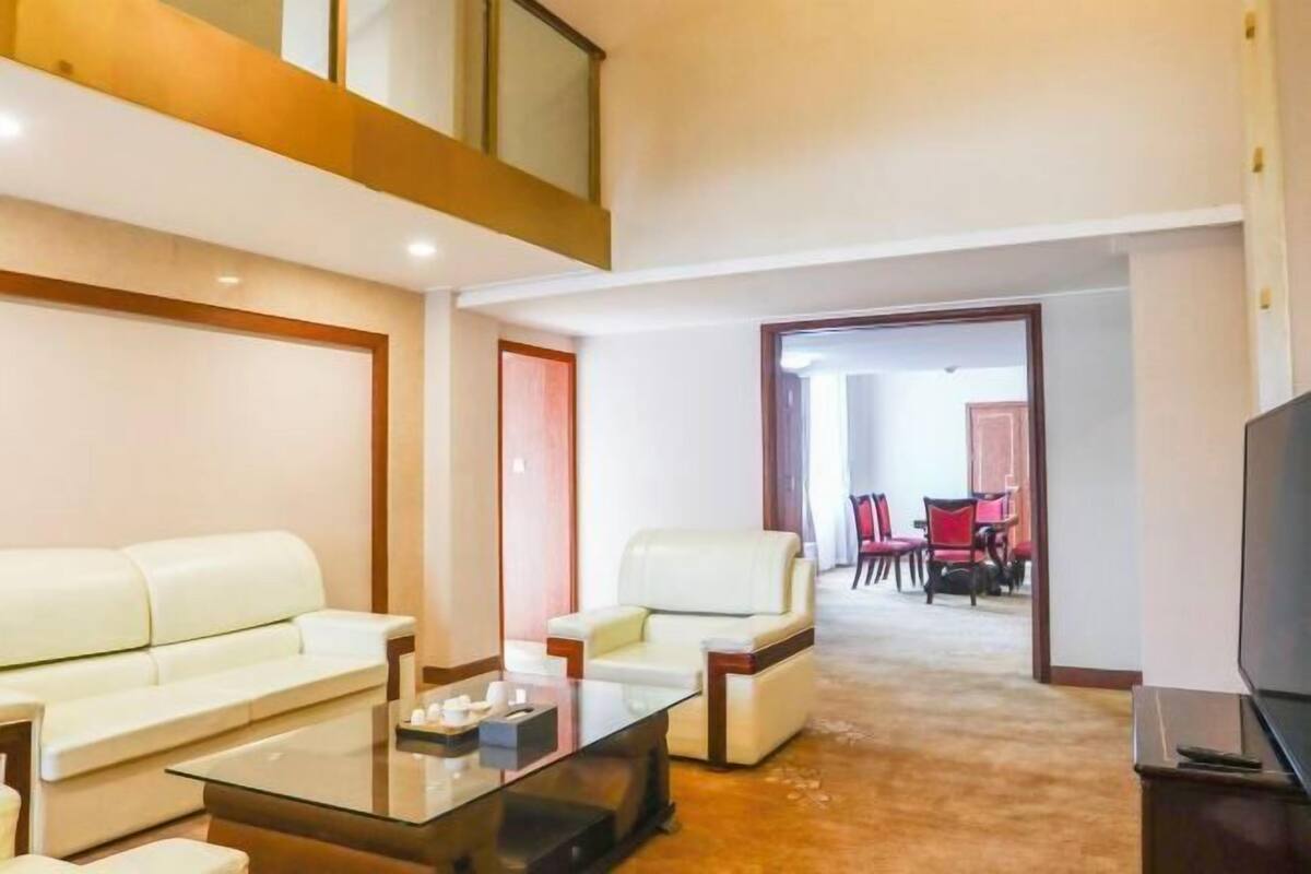 Uma sala de estar com sofá, poltrona e mesa de centro. Há uma televisão à direita e uma área de jantar ao fundo.