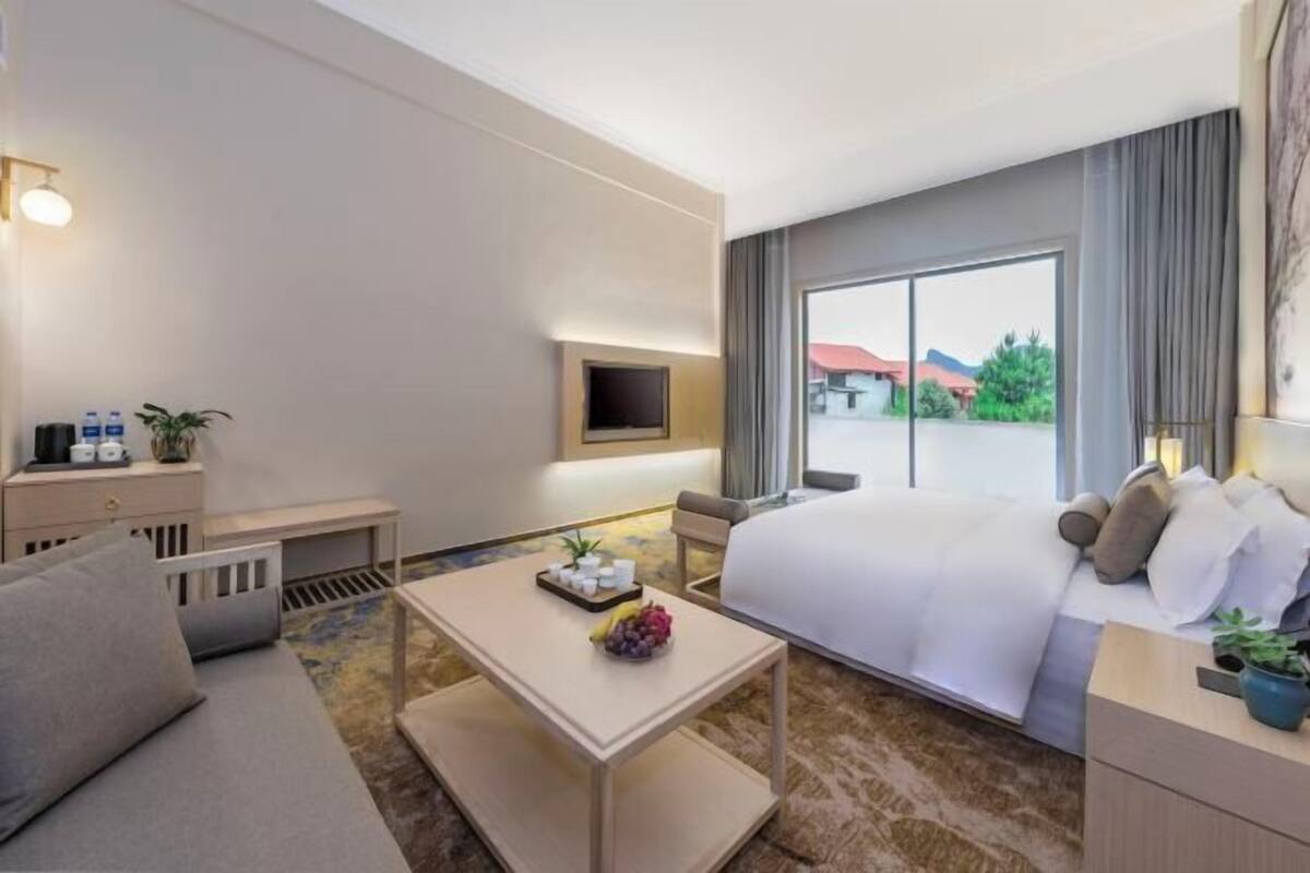 Quarto de hotel com cama, televisão, escrivaninha com planta e janela com vista para edifícios.