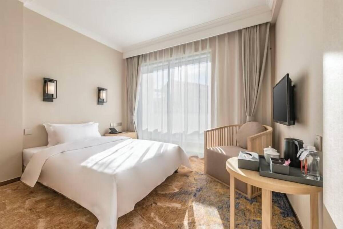 Quarto de hotel com cama, escrivaninha, cadeira, televisão e janela com cortinas.