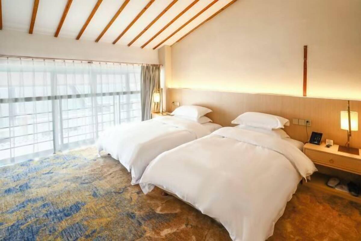 Quarto de hotel com duas camas, uma janela ampla e teto de madeira.