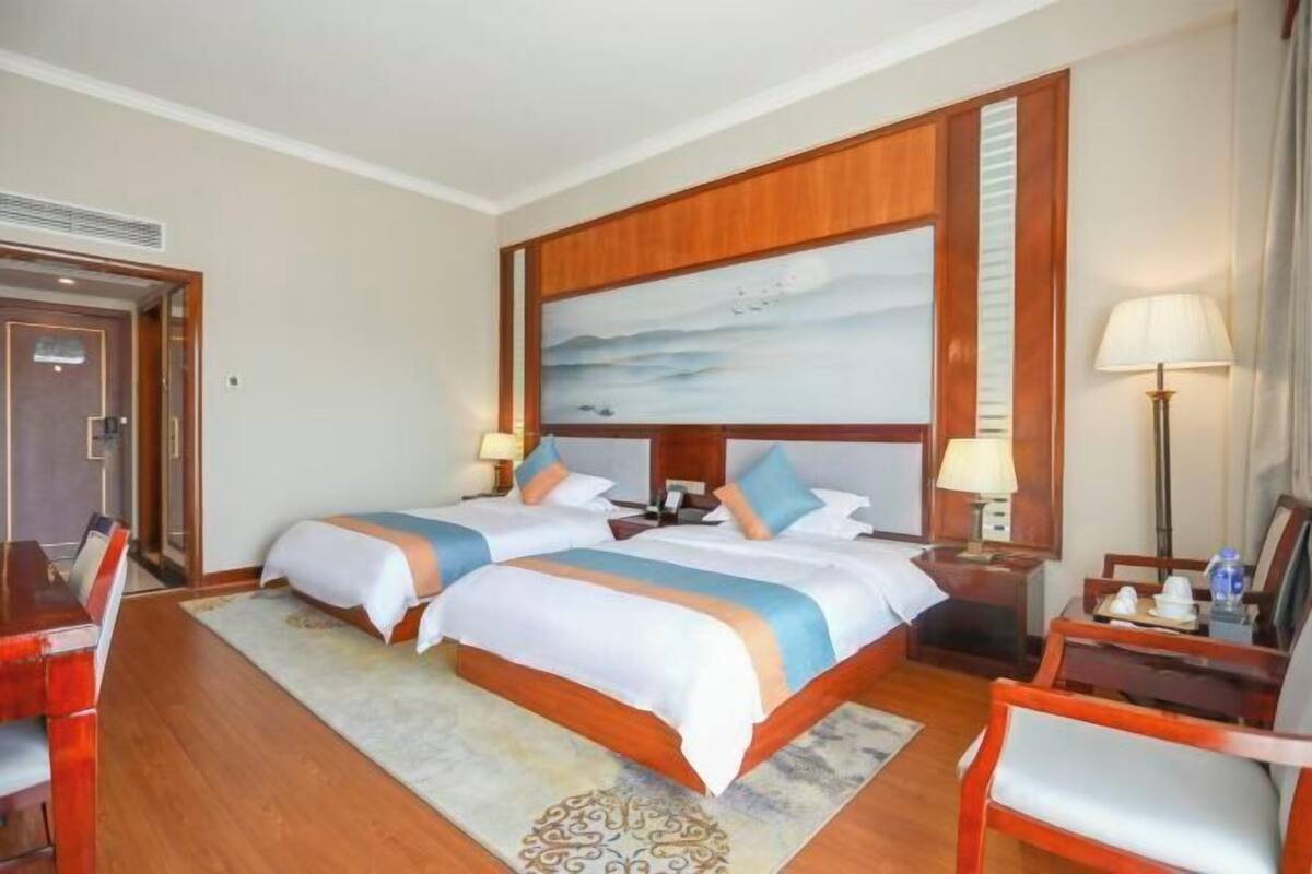 Quarto de hotel com duas camas, uma escrivaninha, uma cadeira, uma mesinha de cabeceira e um quadro na parede.