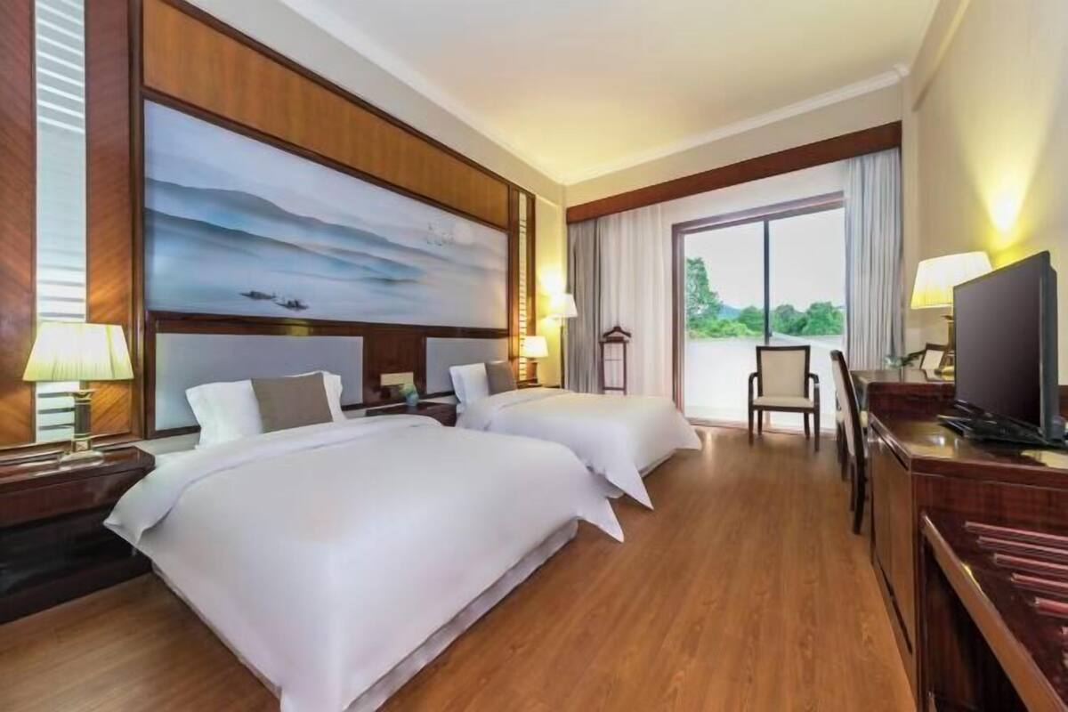 Quarto de hotel com duas camas, um quadro grande, uma televisão e uma escrivaninha com cadeira.