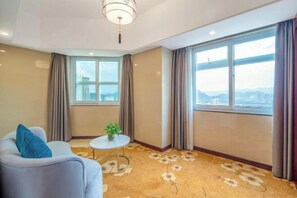 Quarto de hotel moderno com uma janela ampla, uma mesa redonda, um sofá com almofada azul e um tapete floral.